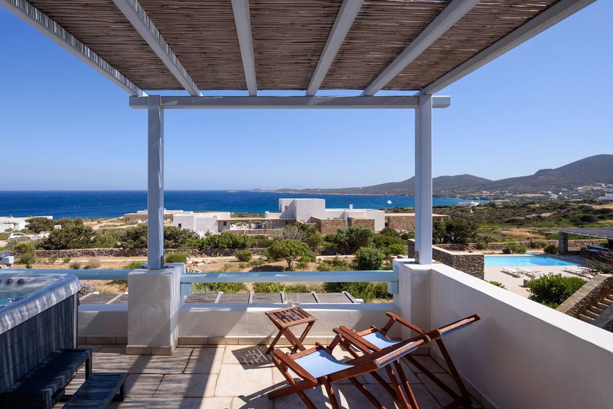 Uma varanda com pérgola de madeira, hidromassagem e espreguiçadeiras de madeira, com vista para uma piscina e o mar.