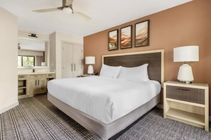 Quarto de hotel com uma cama grande, um ventilador de teto, mesa de cabeceira com abajur, cômoda e janela com cortinas.