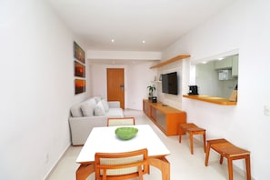 Sala de estar moderna com sofá branco, mesa de jantar com tigela verde, cadeiras de madeira, área de cozinha com TV e armário de madeira.