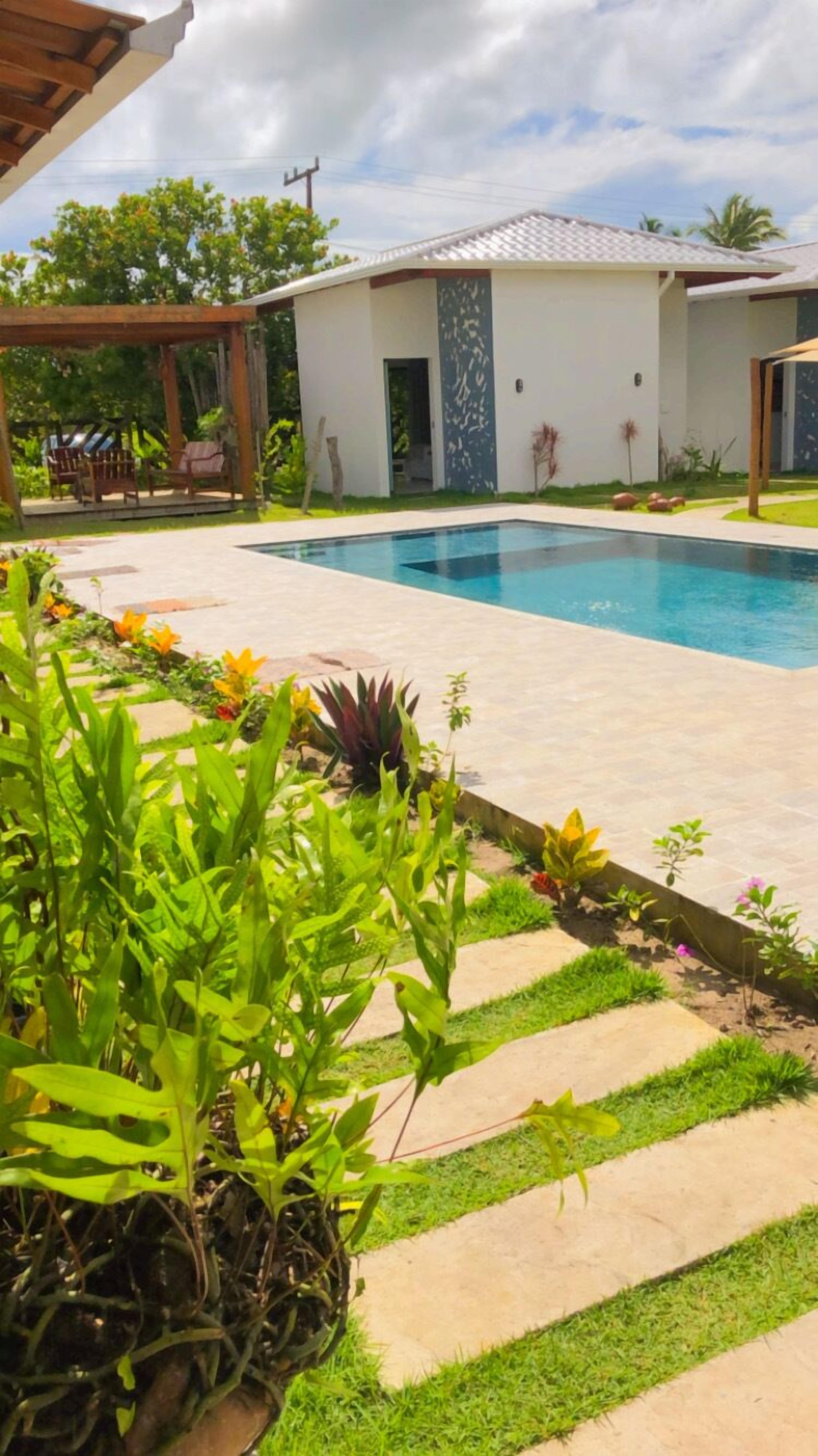 Piscina com deck revestido, rodeada por um jardim com diversas plantas e uma pérgula de madeira.