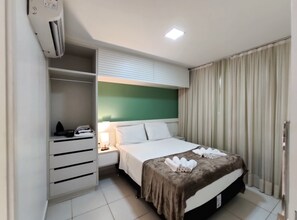 Quarto de hotel com cama, mesa de cabeceira, abajur, guarda-roupa e janela com cortinas.
