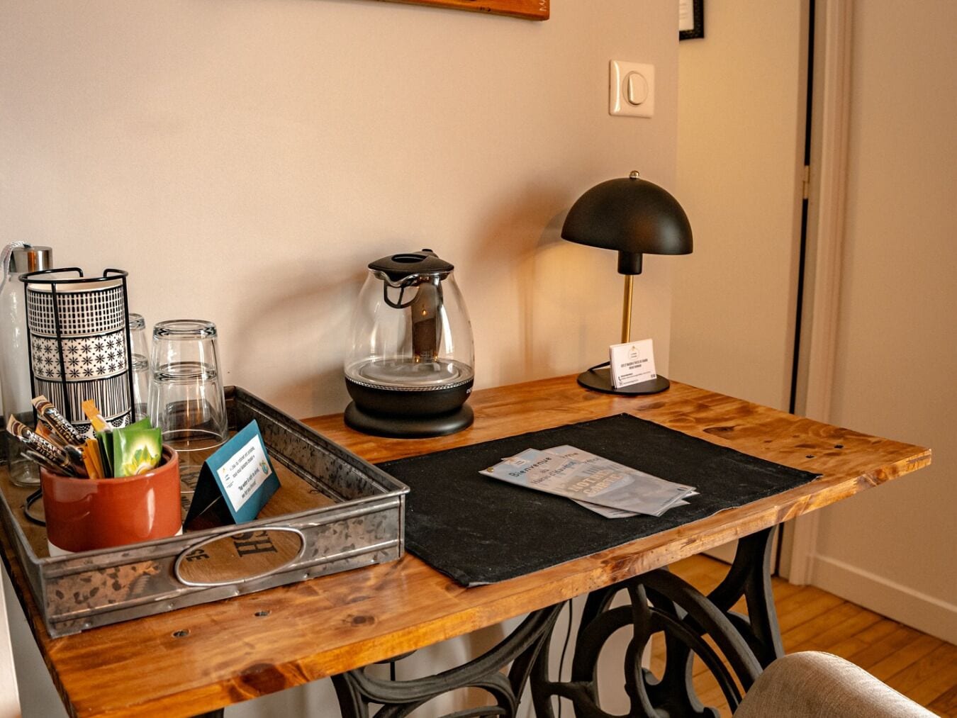 Uma mesa de madeira com um jogo de toalha preto, uma cafeteira, um abajur e uma bandeja com utensílios e um cartão.