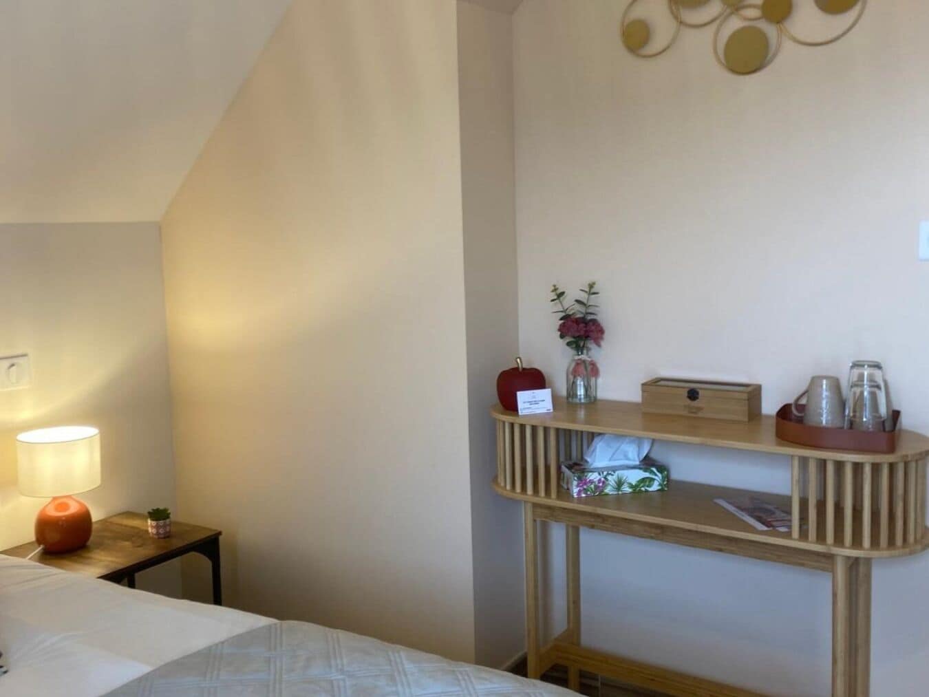 Quarto de hotel com uma cama, uma mesa de cabeceira com abajur, um console de madeira com um vaso de flores, uma maçã vermelha e um tabuleiro com cafeteira e copos.