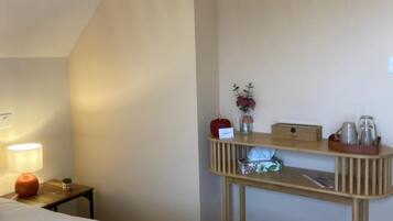Quarto de hotel com uma cama, uma mesa de cabeceira com abajur, um console de madeira com um vaso de flores, uma maçã vermelha e um tabuleiro com cafeteira e copos.