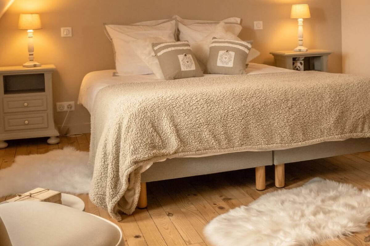 Um quarto com uma cama, duas mesas de cabeceira com luminárias, um criado-mudo, um tapete branco fofo e uma cadeira.