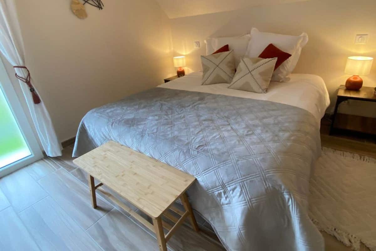 Quarto com cama, almofadas, abajures, um banco de madeira e um ornamento na parede.