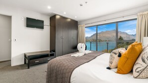 Quarto com cama grande, televisão na parede e vista para montanhas e água através de uma janela ampla.