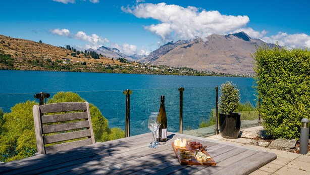 Uma mesa de madeira com uma garrafa de vinho, um copo e uma tábua de queijos, à beira de um lago, com montanhas ao fundo.