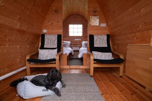Uma cabana acolhedora com duas cadeiras de madeira, uma caminha para um cão pequeno e uma janela que oferece luz natural.