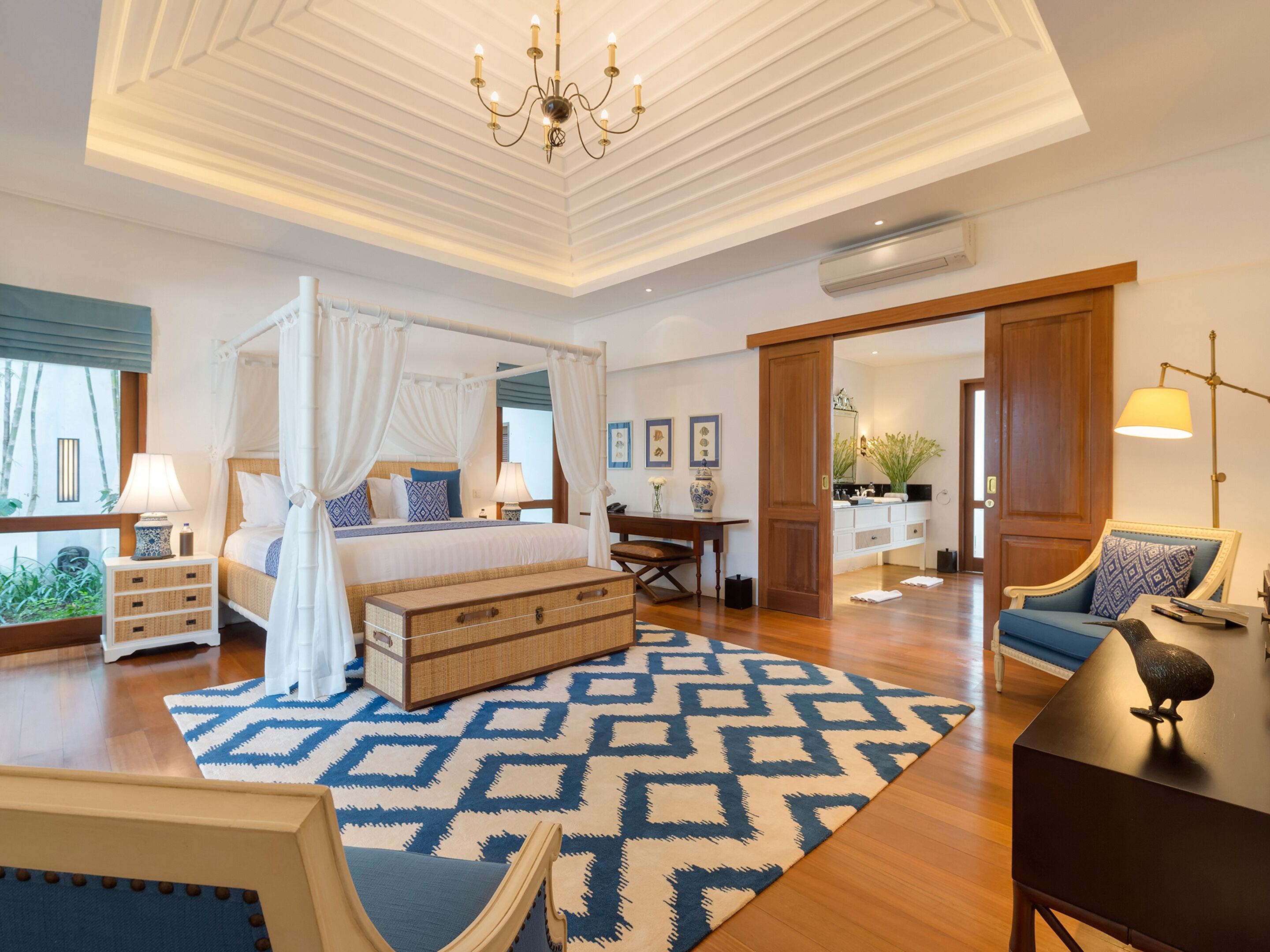 Um quarto espaçoso com cama de dossel, um espaço de estar com uma poltrona e mesa, e um banheiro visível por uma porta aberta.