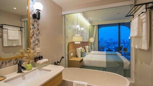 Quarto de hotel moderno com uma banheira grande, uma cama de casal, uma mesinha de vaidade com espelho e vista para a cidade através de uma janela ampla.