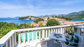 Varanda com mesa e cadeiras, vista para uma vila costeira, com visão clara do mar e montanhas.
