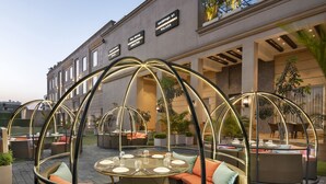 Uma área de refeições ao ar livre com mesas e cadeiras redondas, cercada por um espaço coberto com assentos e vista para um edifício com sinalização.