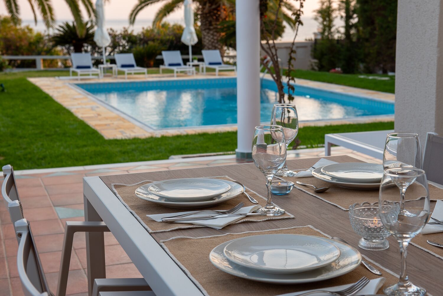 Mesa de jantar posta para dois com vista para a piscina, cadeiras de exterior e céu claro.