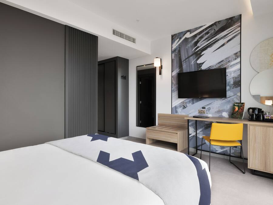 Quarto de hotel moderno com uma cama grande, uma escrivaninha com cadeira, uma TV de tela plana na parede e um espelho amplo.