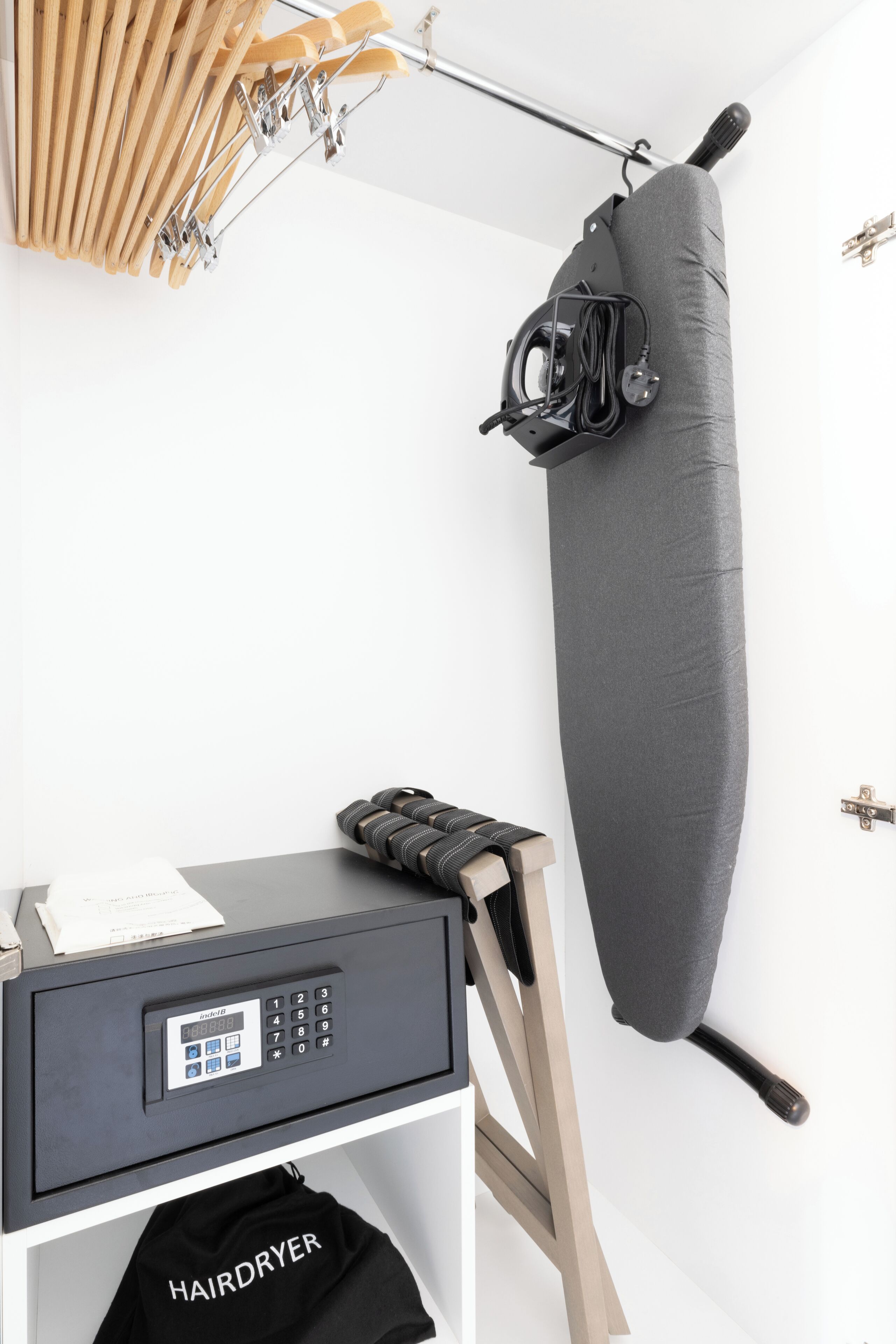Um secador de cabelo em um suporte, uma tábua de passar roupa cinzenta e uma bolsa preta com a etiqueta "HAIRDRYER" em uma prateleira branca.