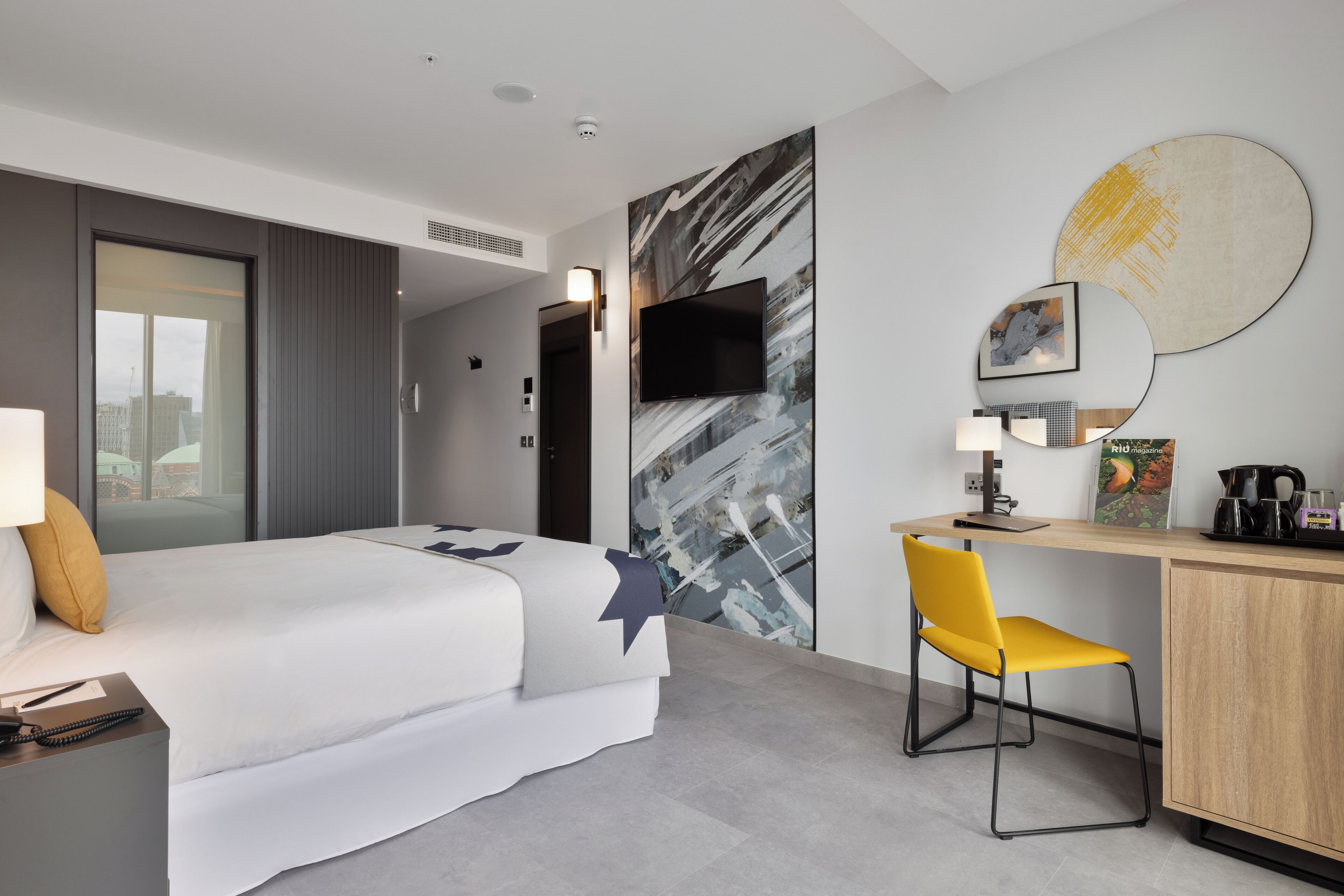 Quarto de hotel moderno com cama, escrivaninha, cadeira e uma grande obra de arte abstrata na parede.