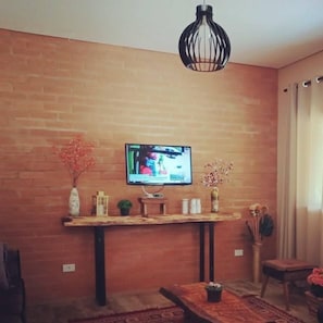Uma sala de estar com parede de tijolos, uma televisão de tela plana fixada nela, uma mesa de console de madeira com objetos decorativos, uma luminária pendente preta e um tapete estampado.