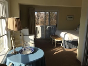 Quarto de hotel com cama, mesa com abajur, cadeira, varanda e vista para árvores, e uma pequena mesa com prato e copos.