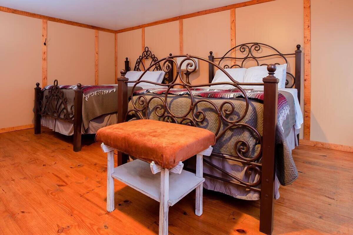 Quarto com duas camas, uma com estrutura de metal e outra de madeira. Há um banco em frente às camas.