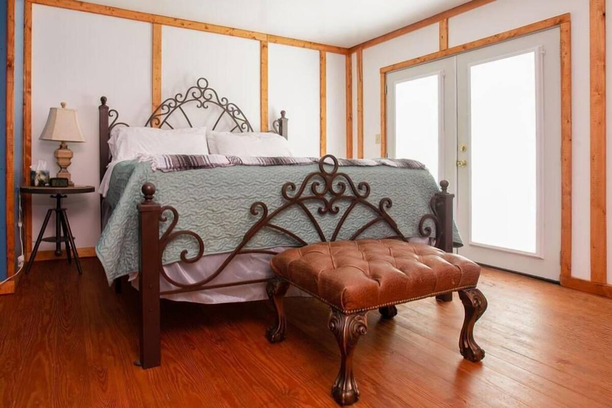 Quarto com estrutura de cama de madeira, colcha verde, banquinho estofado marrom e mesa de cabeceira com luminária.