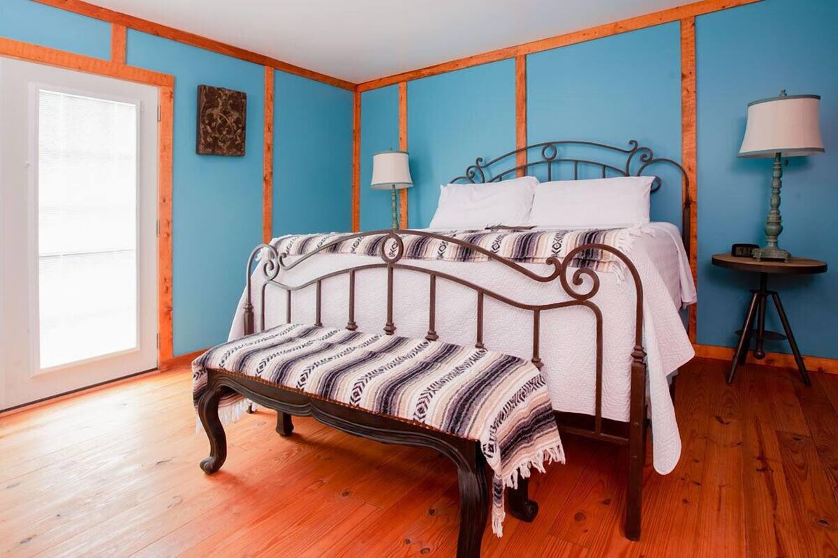 Quarto com piso de madeira, estrutura de cama de metal, um banco, duas mesas de cabeceira e uma janela.