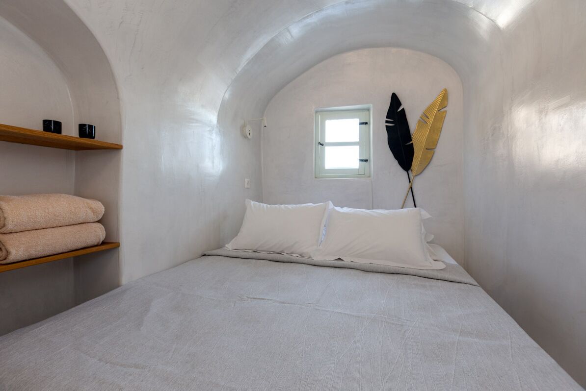 Quarto pequeno com arco, cama, almofadas, janela e prateleira com toalhas.