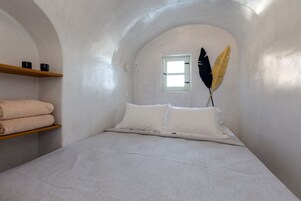 Quarto pequeno com arco, cama, almofadas, janela e prateleira com toalhas.