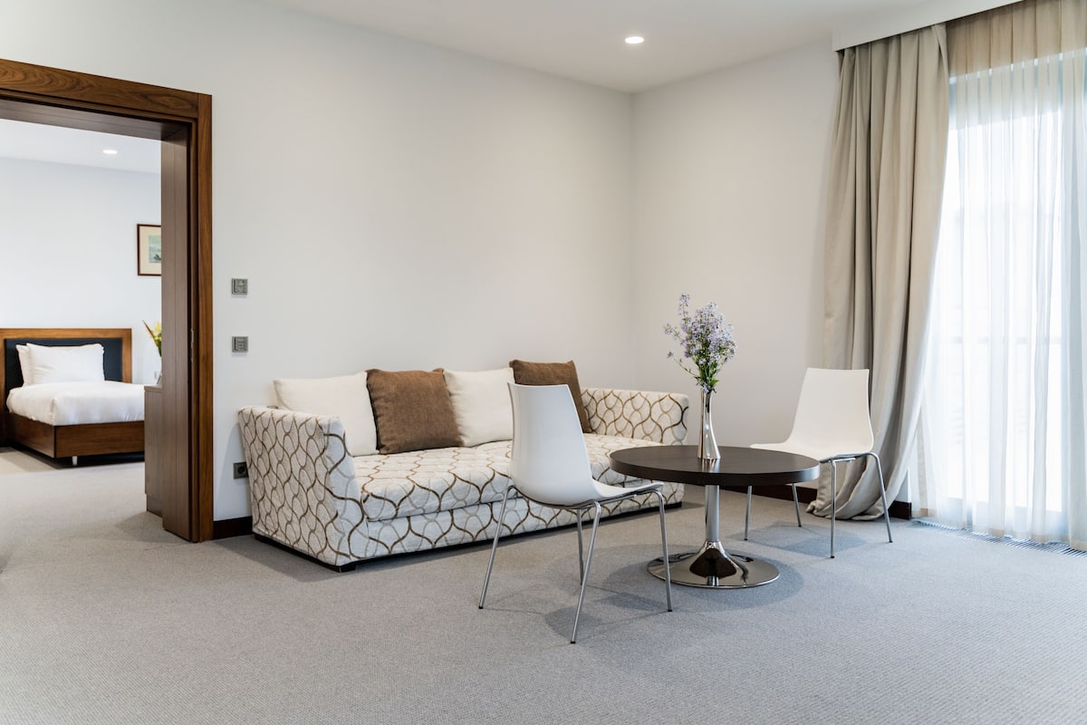Quarto de hotel moderno com sofá, mesa redonda e uma cadeira. Uma cama aparece ao fundo e uma janela com cortinas.