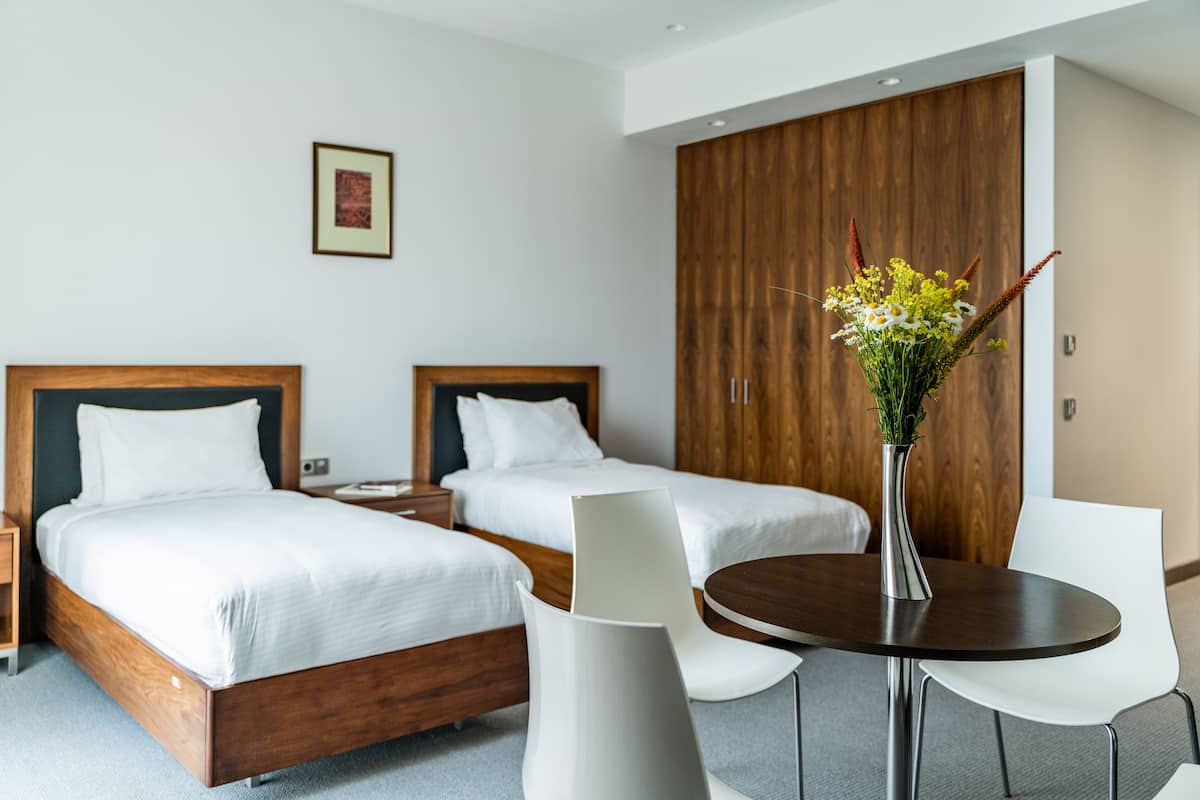 Quarto de hotel com duas camas, um guarda-roupa de madeira, uma mesa redonda com um vaso de flores e uma cadeira branca.