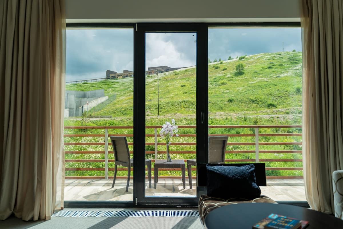 Quarto com uma grande porta de vidro deslizante que dá para uma varanda com vista para uma colina gramada e edifícios ao longe. Há uma cama com um travesseiro e um livro sobre a mesa.
