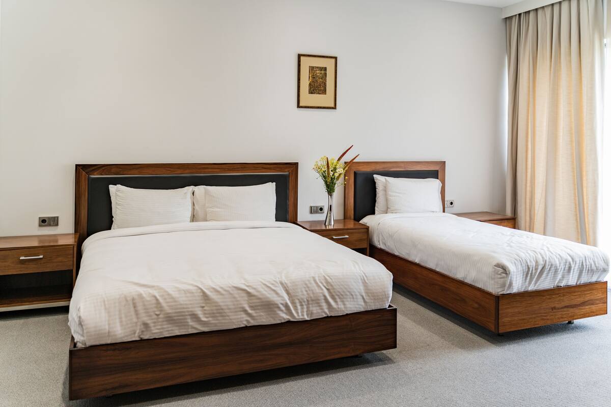 Quarto de hotel com duas camas de madeira, uma mesa de cabeceira, um criado-mudo, um quadro na parede e uma janela com cortinas.