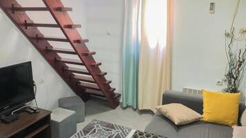Uma sala de estar com escada de madeira, um sofá cinza com almofadas amarelas, uma televisão de tela plana e um tapete estampado.