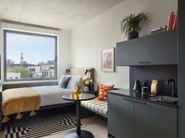 Quarto de hotel compacto com cama, uma pequena mesa de jantar, cozinha compacta e uma grande janela com vista para a cidade.