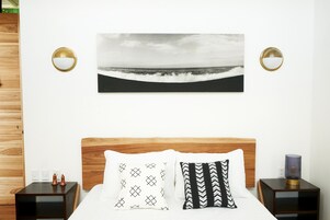 Quarto com cabeceira de madeira, cama com almofadas estampadas, mesa de cabeceira com luminária e um quadro pendurado na parede retratando uma cena de praia.