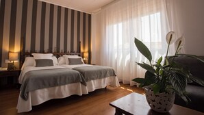 Quarto de hotel com duas camas, uma janela ampla com cortinas leves, piso de madeira e uma planta em vaso sobre uma mesa.