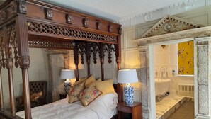 Uma cama de dossel com esculturas elaboradas, duas mesas de cabeceira com luminárias, e vista para um banheiro com papel de parede floral amarelo.