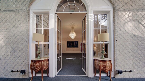 Uma porta em arco com duas mesas laterais, cada uma com uma luminária, que dá acesso a um quarto com um candelabro.