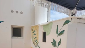 Um pátio iluminado pelo sol, com piso de azulejos, plantas em vasos, uma pequena janela e uma parede decorada com motivos de folhas.