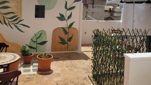 Um terraço no telhado com mesa e cadeiras, plantas em vasos, um painel decorativo com motivos de folhas e um guarda-sol azul.