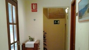 Um corredor estreito com uma porta de madeira à esquerda, uma pequena mesa com uma planta e uma placa indicando uma saída.