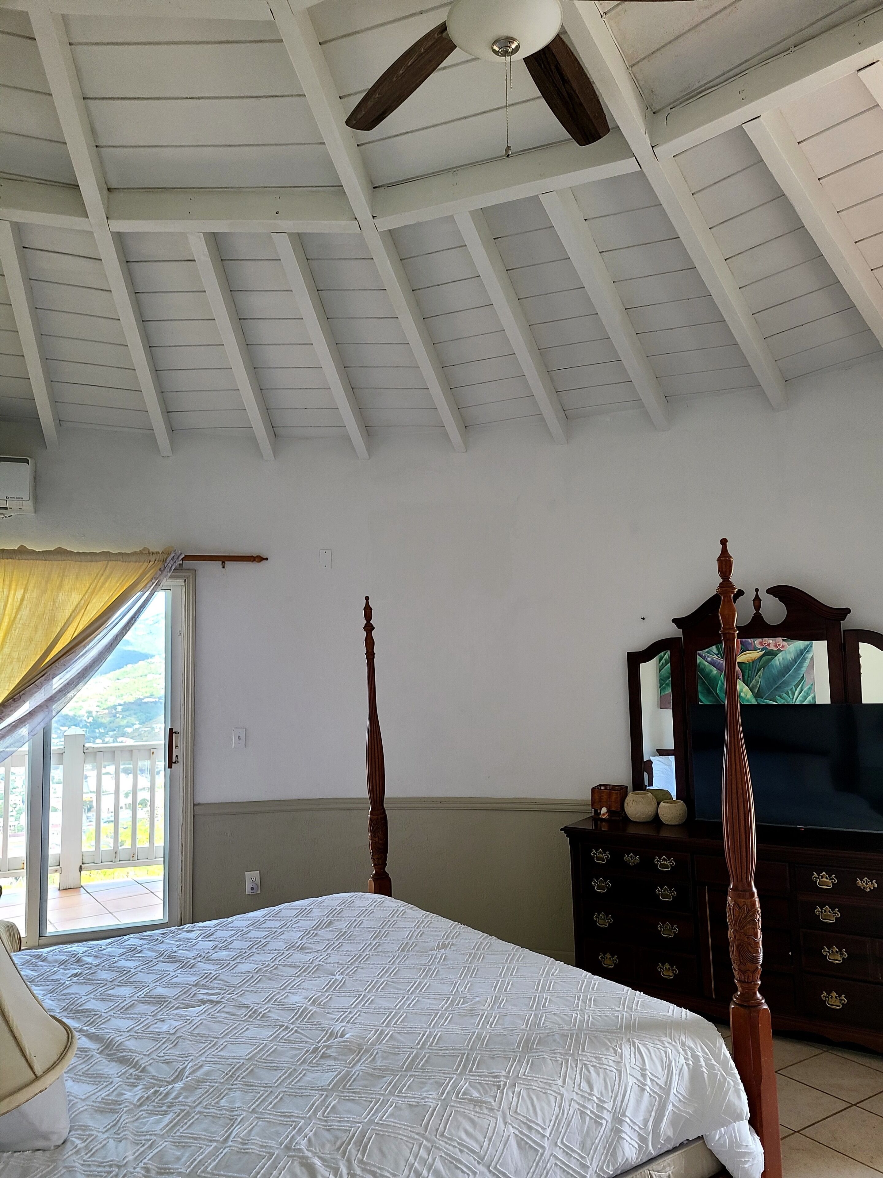 Quarto com cama de dossel, ventoinha no teto, cômoda de madeira e uma janela grande com vista para o verde.