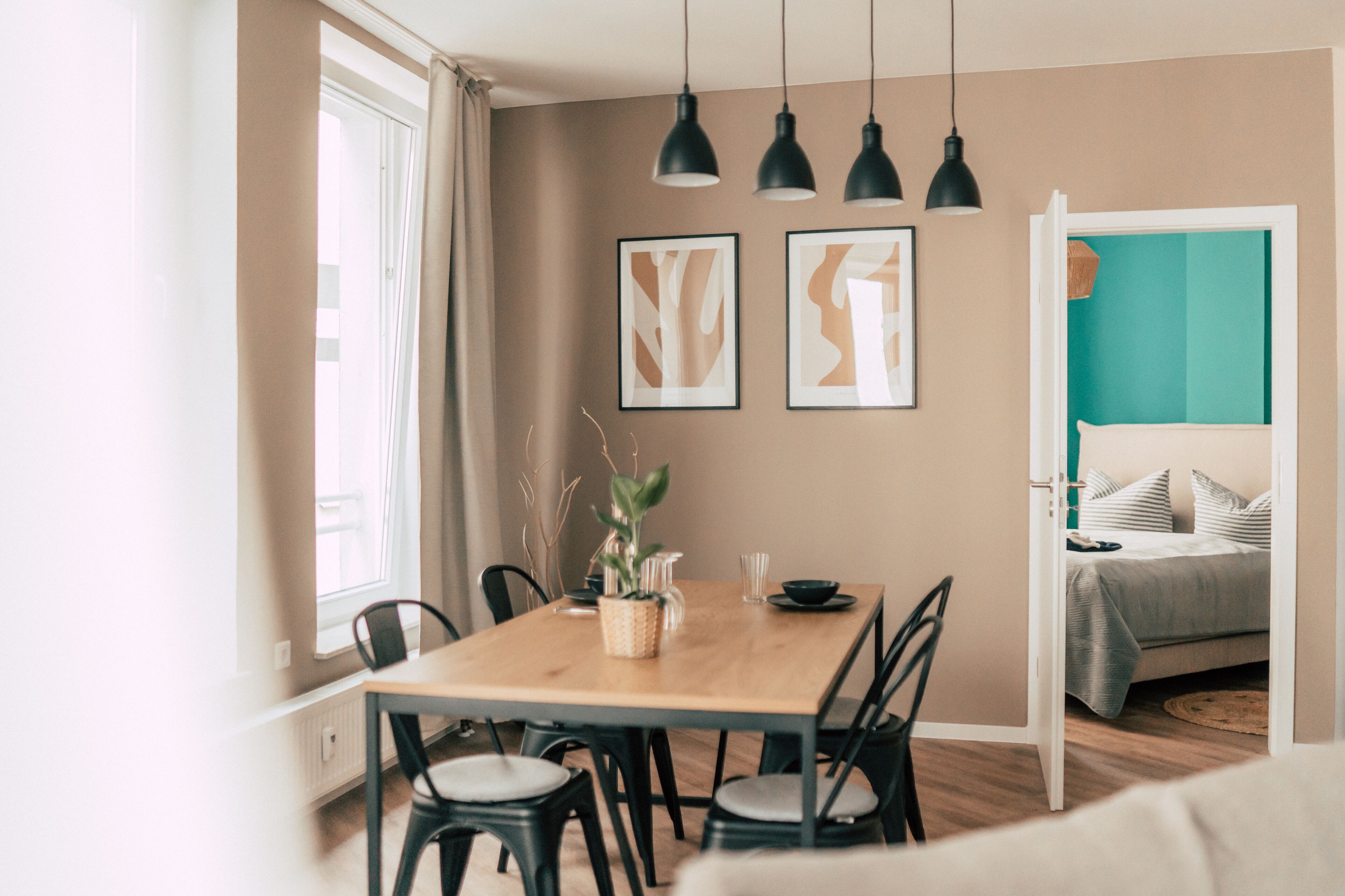 Uma área de refeições com mesa de madeira, cadeiras pretas e luminárias pendentes. Há obras de arte abstratas emolduradas na parede e uma planta sobre a mesa.