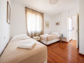 Quarto de hotel com duas camas, uma mesinha de cabeceira, uma cadeira e uma porta que dá para outro cômodo.