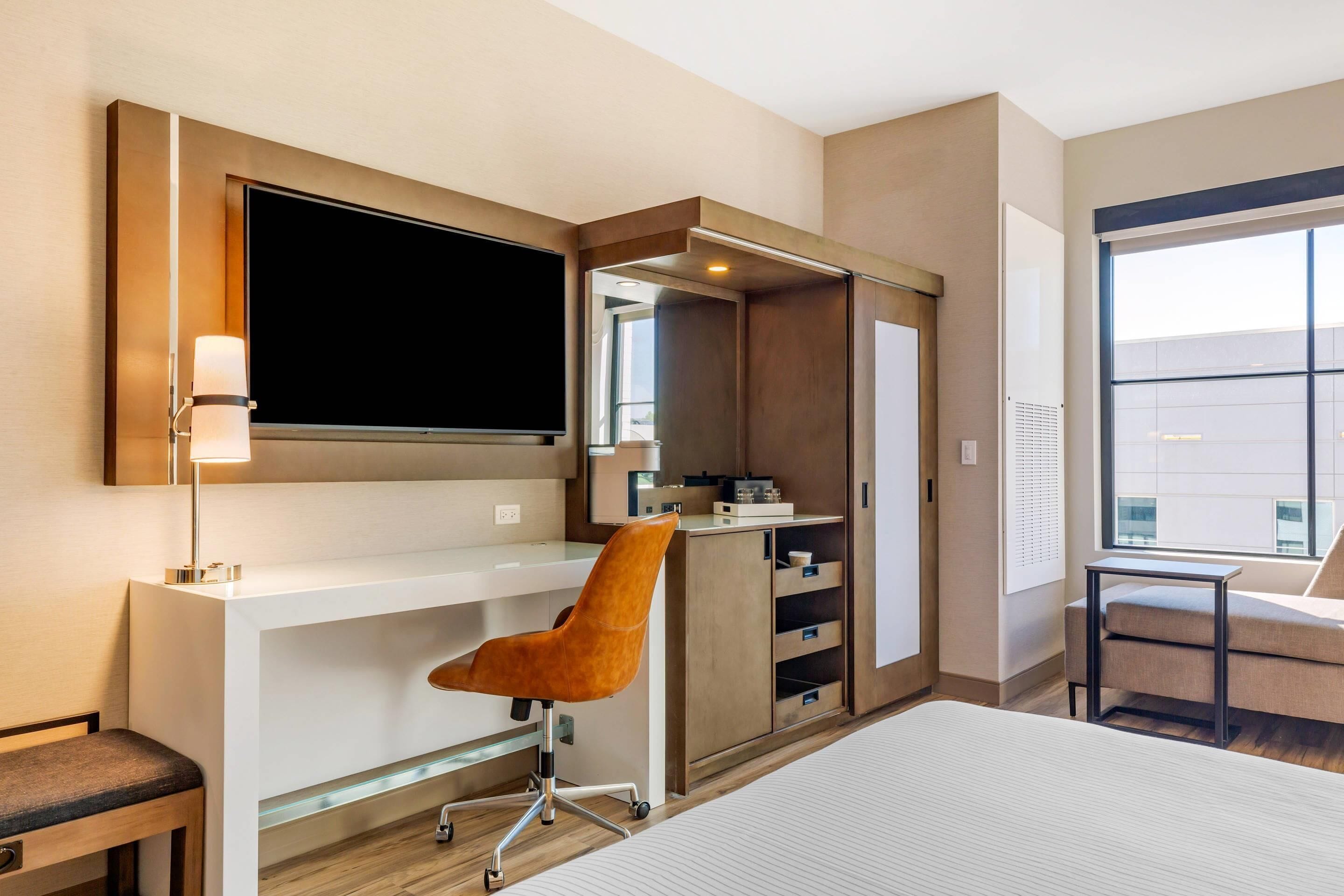 Quarto de hotel moderno com uma cama grande, uma escrivaninha com cadeira, TV de tela plana, minibar e uma janela ampla com vista.