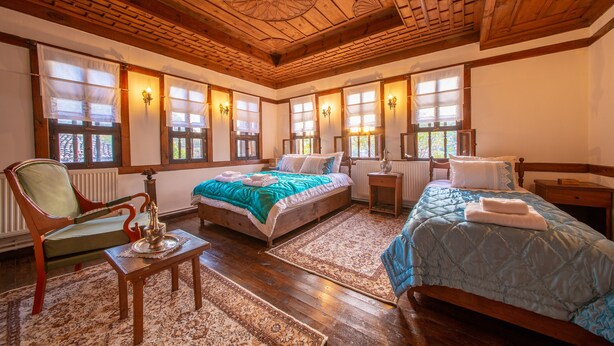 Um quarto espaçoso com piso de madeira, uma cama grande, uma cama menor, uma cadeira e uma mesa com um objeto decorativo.