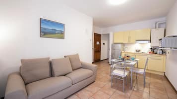 Uma sala de estar com sofá e uma mesa de jantar com cadeiras. Há uma área de cozinha com armários e eletrodomésticos.