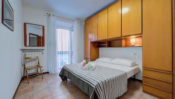 Quarto com cama, guarda-roupa de madeira, radiador, uma pequena cadeira de madeira e janela com cortinas.