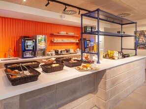 Buffet de café da manhã com diversos alimentos, máquinas de café e uma estante azul.