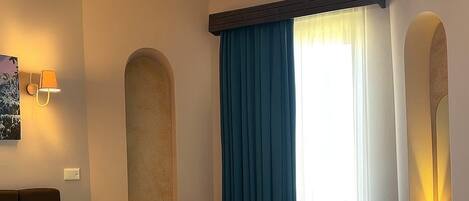 Quarto de hotel com uma cama grande, um ventilador de teto, uma janela com cortinas azuis, uma luminária fixada na parede e um radiador.